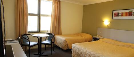Quarto de hotel com duas camas, uma escrivaninha com cadeira, uma mesinha e uma janela com cortinas.