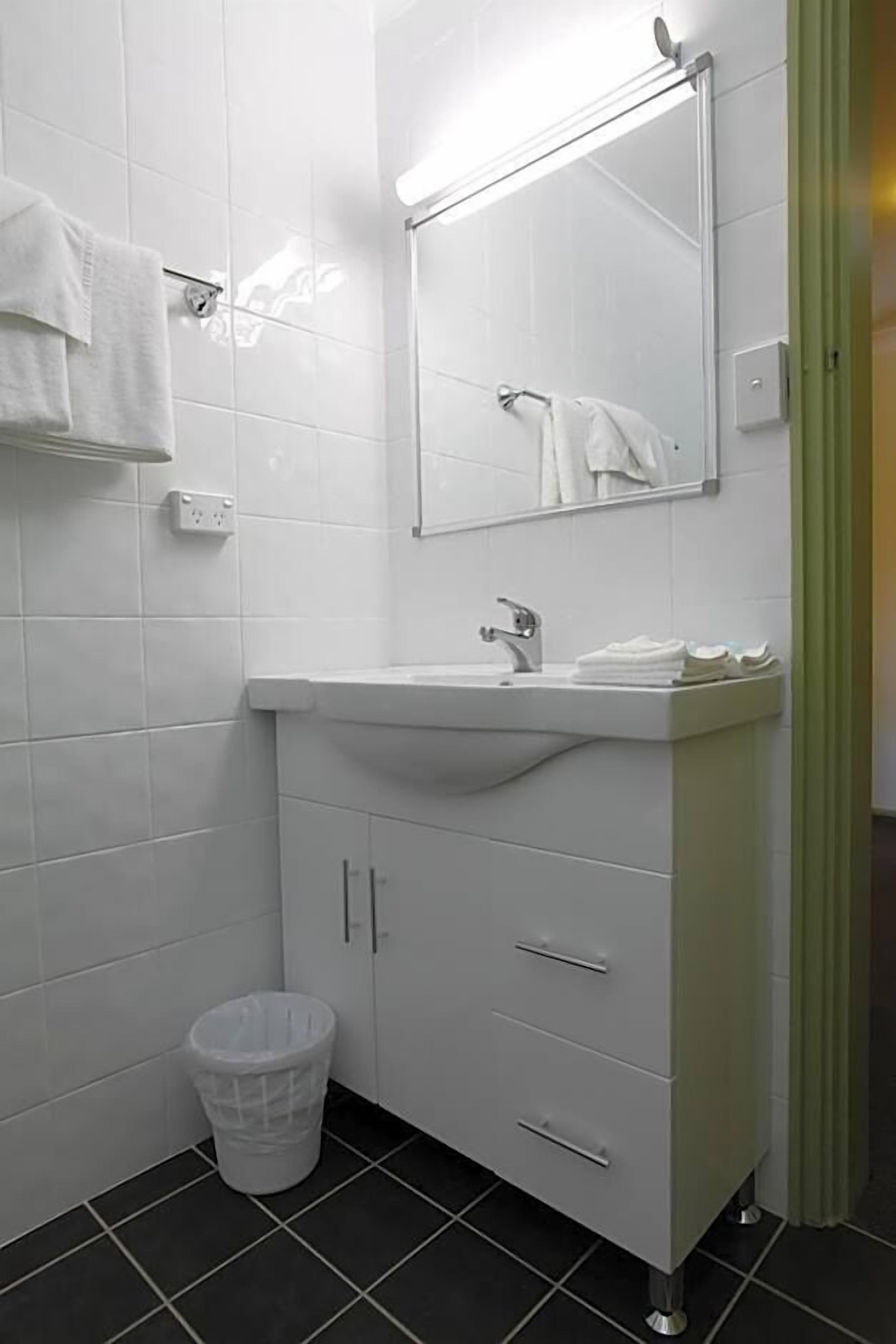Um banheiro com azulejos brancos, pia, espelho, porta-toalhas e um cesto de lixo.