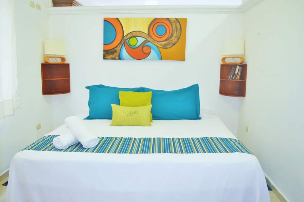 Quarto de hotel com cama, almofadas, colcha listrada, um quadro na parede e duas mesas de cabeceira com luminárias.