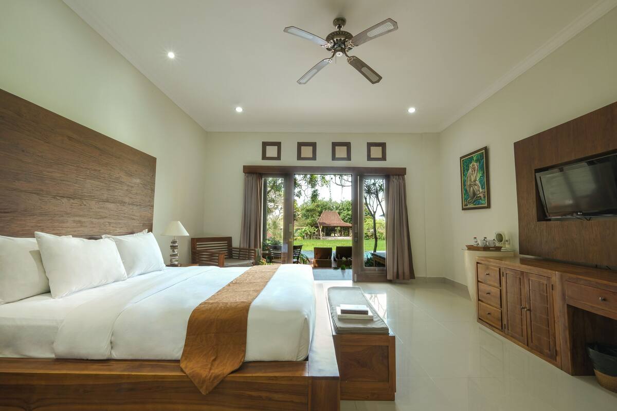 Quarto espaçoso com uma cama grande, cabeceira de madeira, ventilador de teto, televisão e vista para o exterior através de amplas janelas.