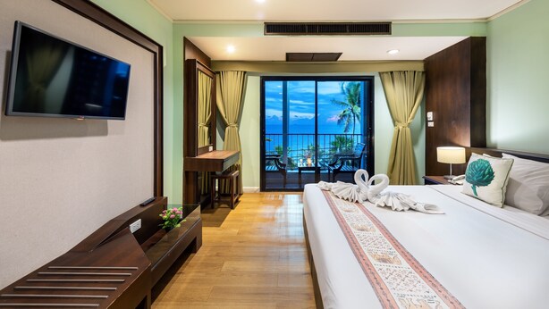 Quarto de hotel com uma cama grande, escrivaninha, televisão e varanda com vista para palmeiras e o céu.