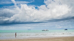 Uma praia com uma pessoa caminhando, barcos na água e um céu nublado.