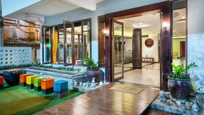 Um hall de hotel com piso de madeira, assentos coloridos, plantas em vasos e uma área de recepção com um aviso.