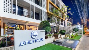 Exterior moderno de um hotel com uma placa indicando "JOMTIEN TWELVE", um jardim bem cuidado com grama sintética e um edifício de vários andares com varandas.