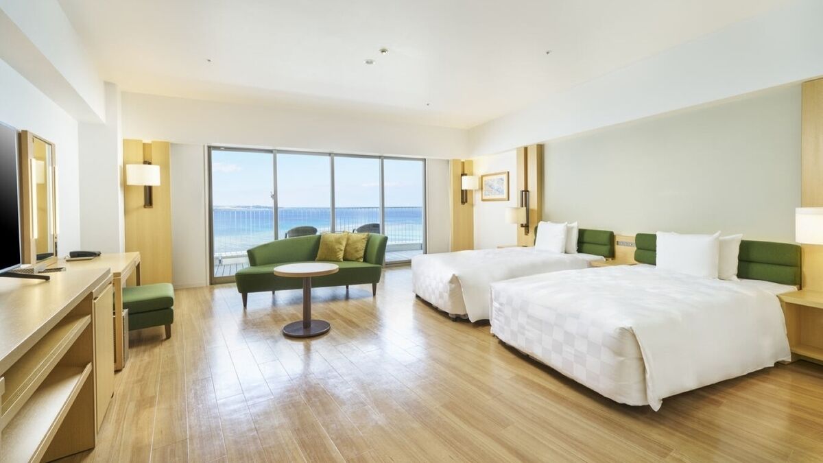 Quarto de hotel com uma janela grande, duas camas, uma escrivaninha e uma poltrona verde.