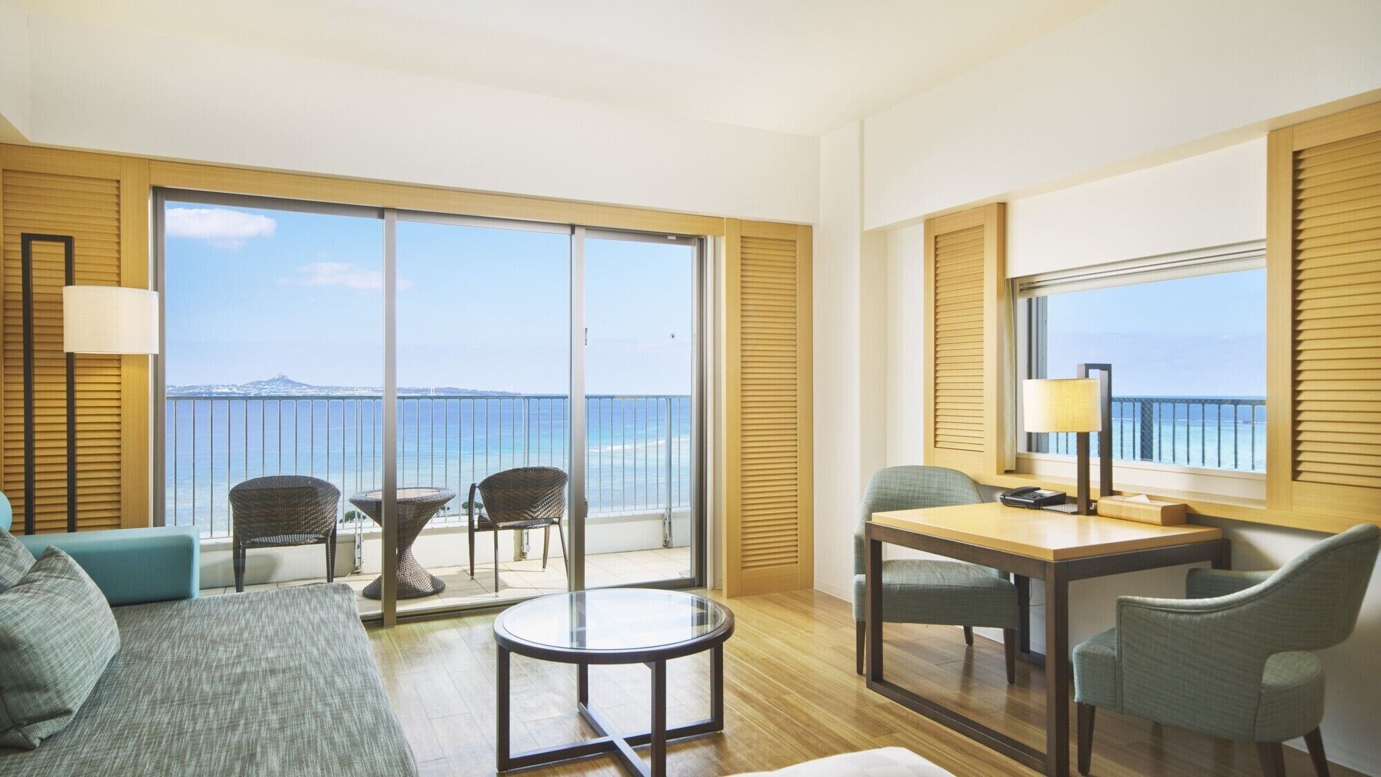 Quarto de hotel com varanda, uma mesa redonda, duas cadeiras, um sofá e um abajur.
