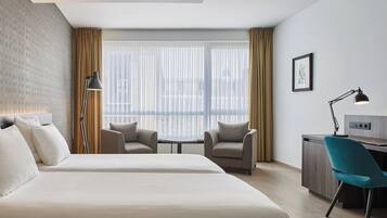 Quarto de hotel com duas camas, uma escrivaninha, uma cadeira, uma luminária e vista para a cidade.