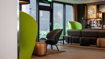 Um lobby moderno com um sofá de couro preto, uma poltrona verde e um banquinho marrom. Apresenta uma parede de tijolos e uma grande janela com vista para o verde.