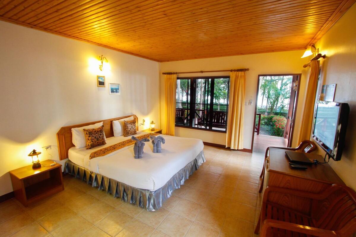 Quarto de hotel com uma cama grande, uma escrivaninha com computador, televisão e varanda com cortinas.