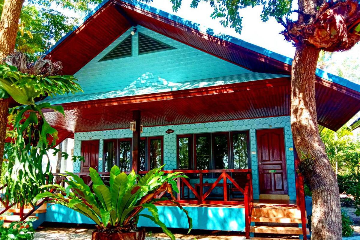 Uma casa de dois andares com telhado vermelho e paredes azuis, rodeada por árvores e plantas.