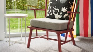 Uma cadeira com almofada estampada, um abajur branco e vista para uma sebe verde através de uma janela.
