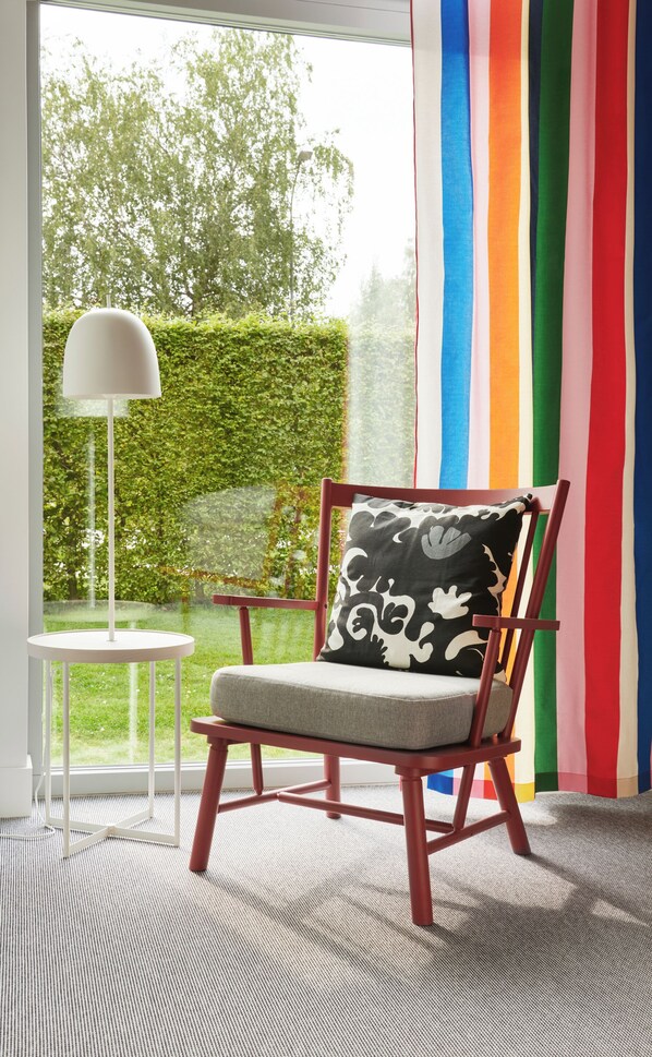 Uma cadeira com almofada estampada, um abajur branco e vista para uma sebe verde através de uma janela.