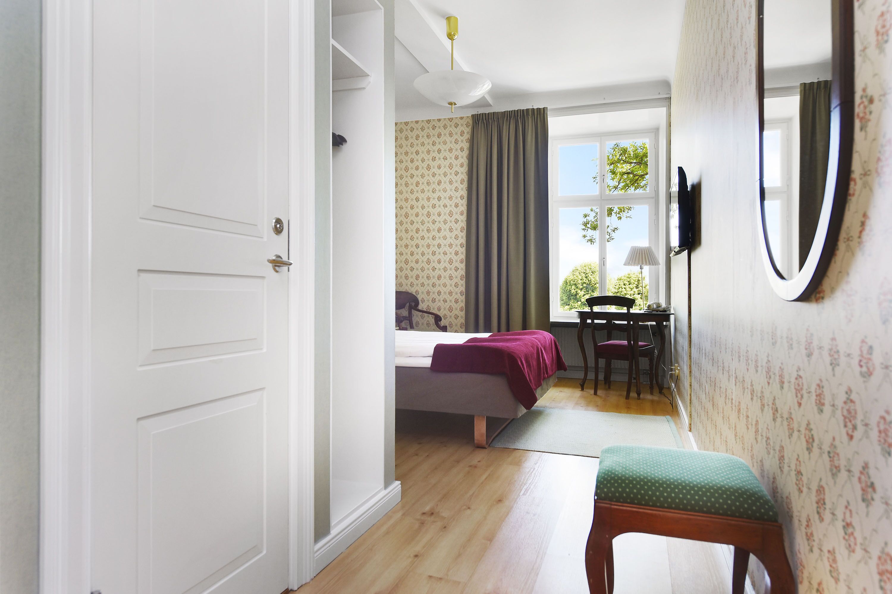 Quarto com cama, escrivaninha e cadeira. Há uma janela com cortinas, um espelho e uma porta.