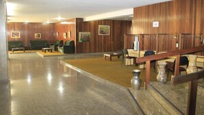 Um saguão com painéis de madeira, balcão de recepção, várias cadeiras, uma escada e um vaso no chão.