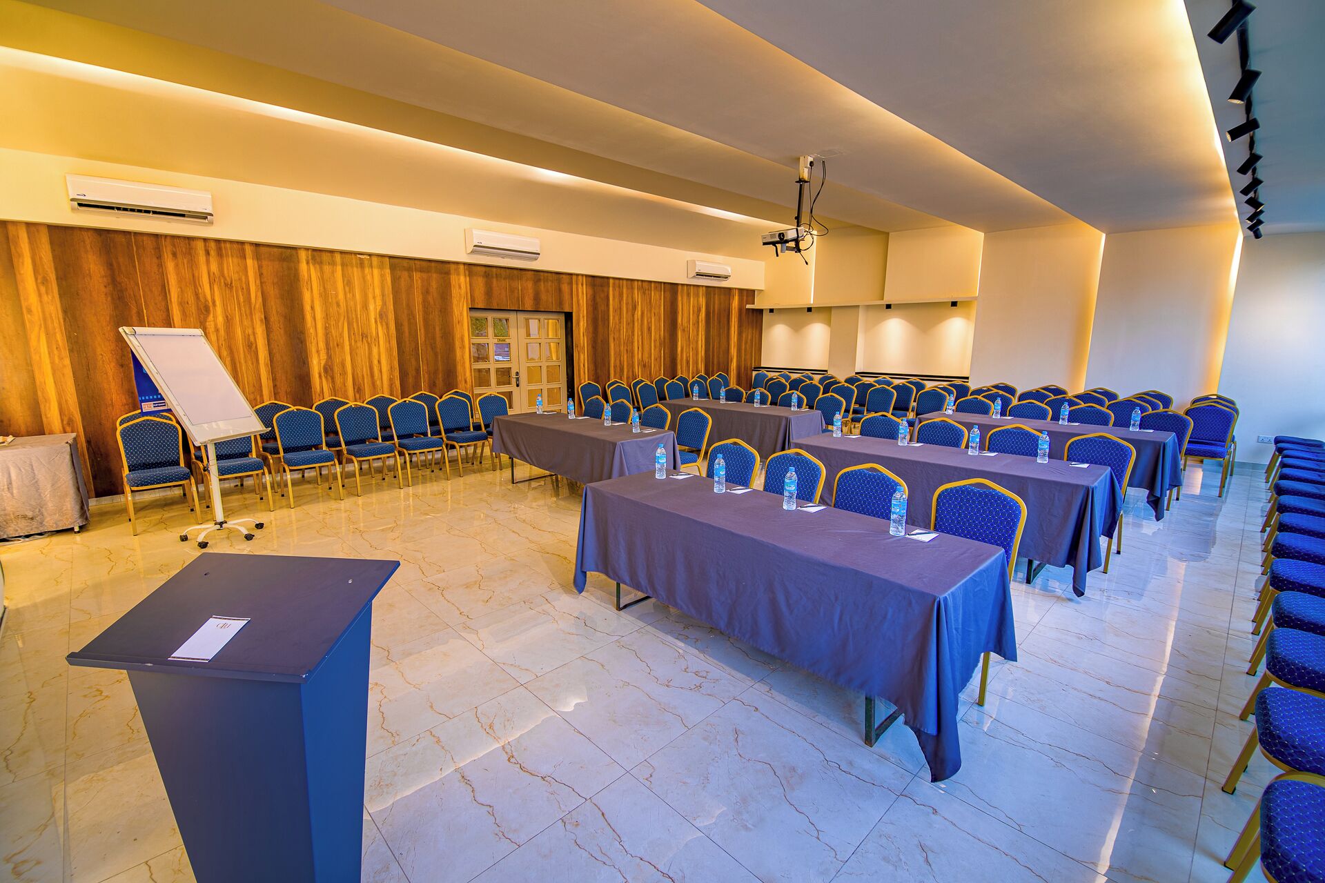 Uma sala de conferências com fileiras de cadeiras azuis, mesas com garrafas de água, um pódio e uma lousa.