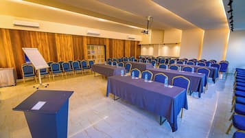 Uma sala de conferências com fileiras de cadeiras azuis, mesas com garrafas de água, um pódio e uma lousa.
