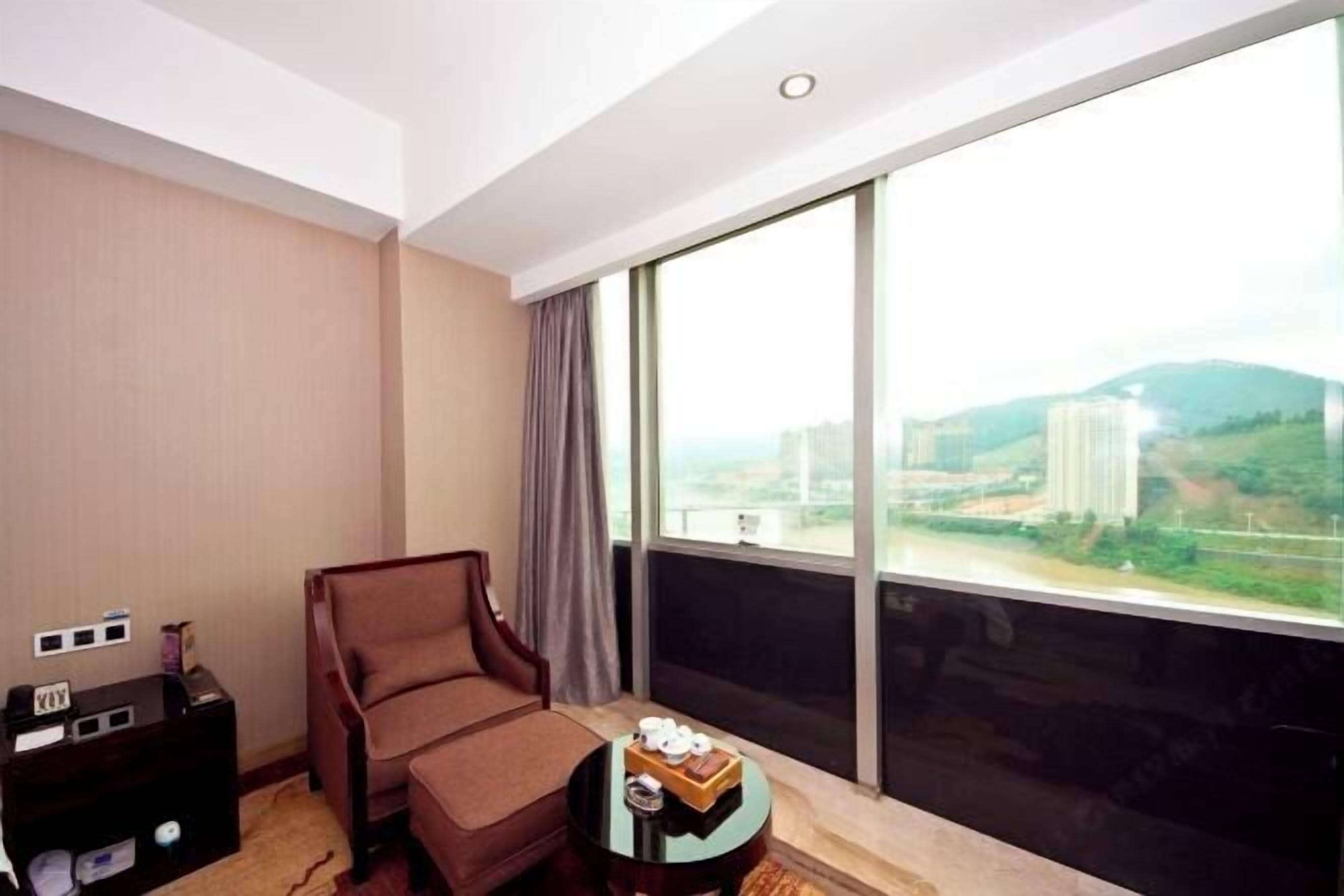 Quarto de hotel com uma janela grande, um sofá, uma mesa pequena com bandeja e uma mesinha de cabeceira escura.