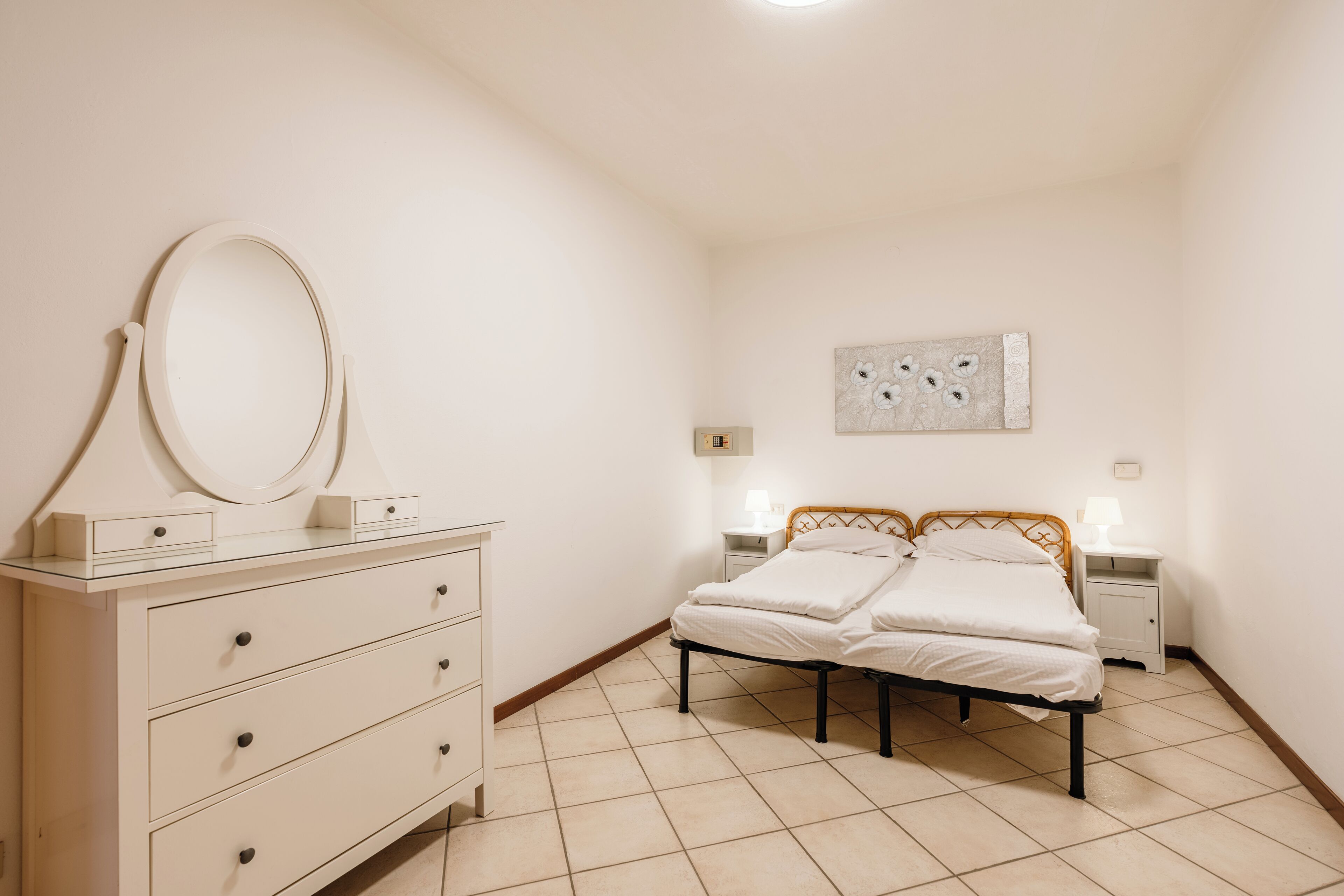 Quarto com cama, cômoda, espelho, mesa de cabeceira e luminária fixada na parede.