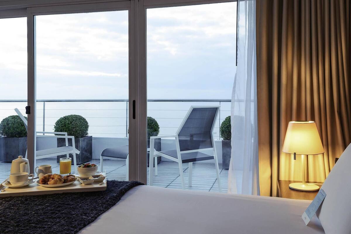 Quarto de hotel com varanda, uma cama, um tabuleiro com itens de pequeno-almoço, uma cadeira e um candeeiro.