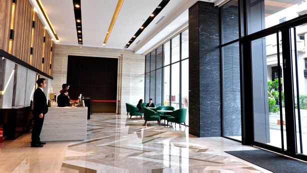 Um hall de hotel moderno com balcão de recepção, área de estar com assentos verdes e grandes janelas com vista para o exterior.