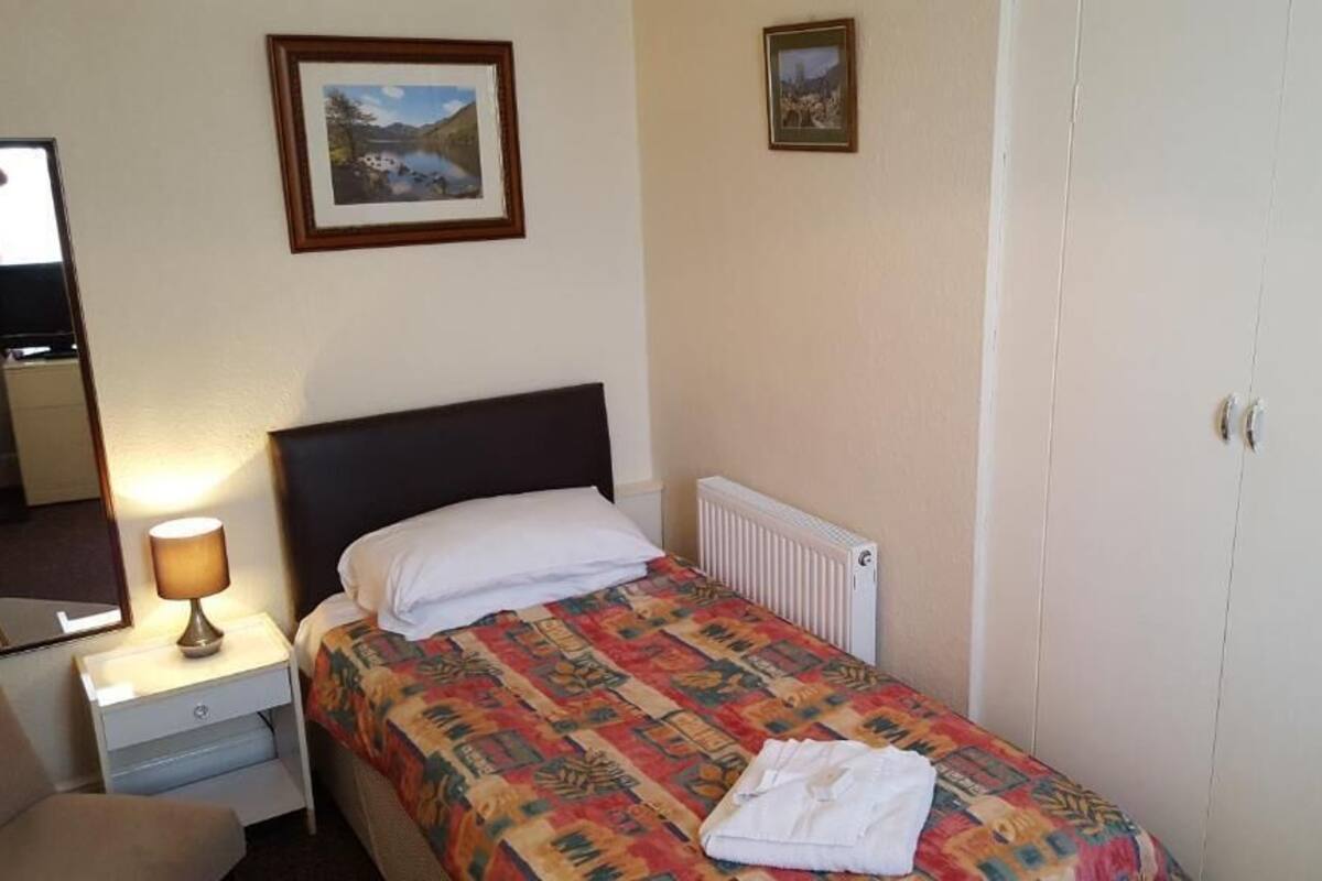 Quarto de hotel com uma cama, uma mesa de cabeceira com abajur, um radiador e duas fotografias emolduradas na parede.