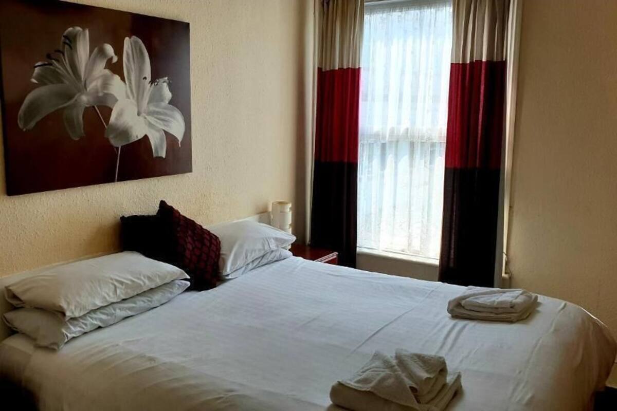 Quarto de hotel com uma cama grande, roupa de cama branca, um quadro de flores na parede e uma janela com cortinas vermelhas e brancas.