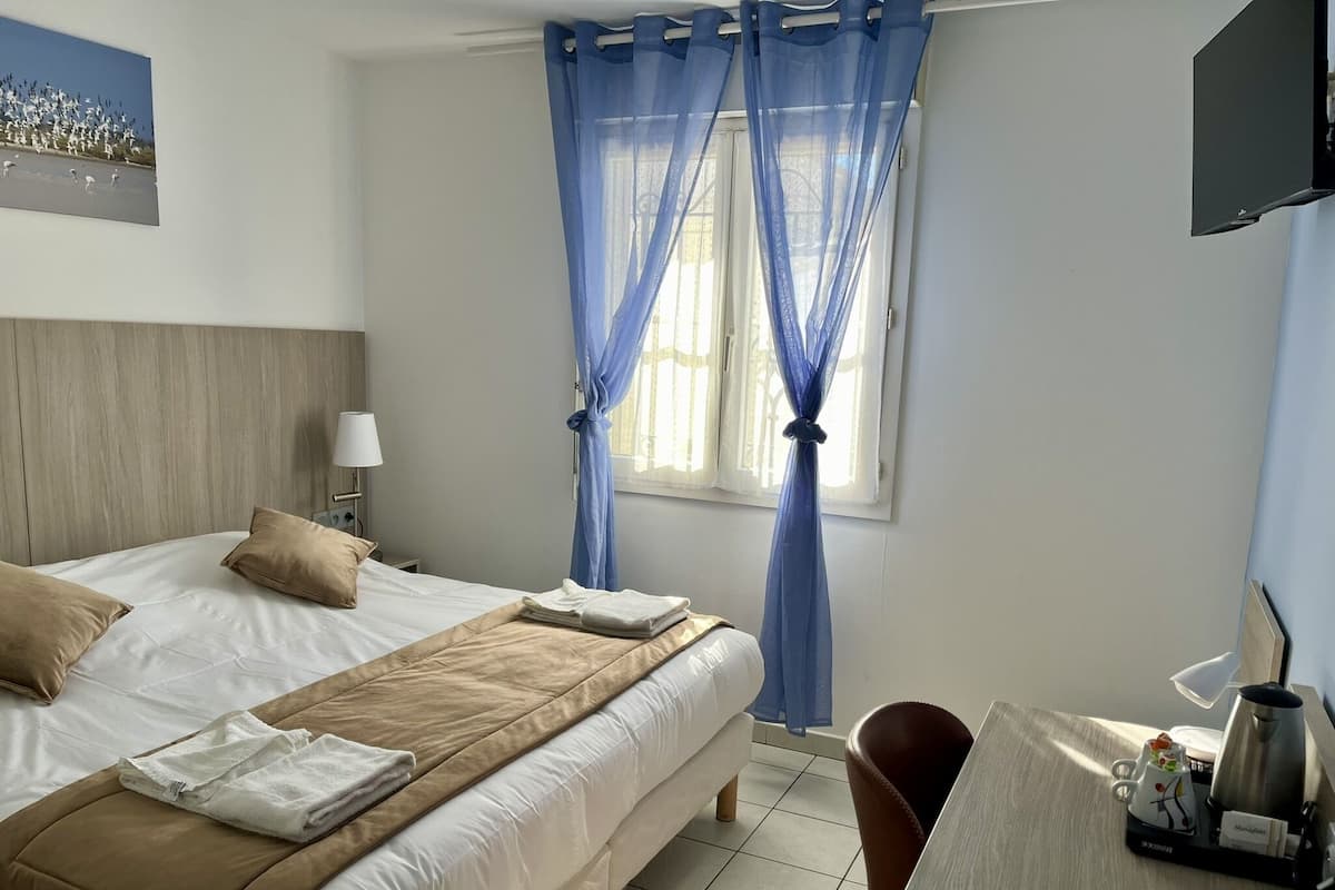 Quarto de hotel com cama, escrivaninha, cadeira, televisão e janela com cortinas azuis.