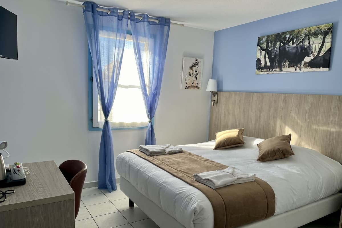 Quarto de hotel com cama, escrivaninha, cadeira, janela com cortinas azuis e luminária na parede.