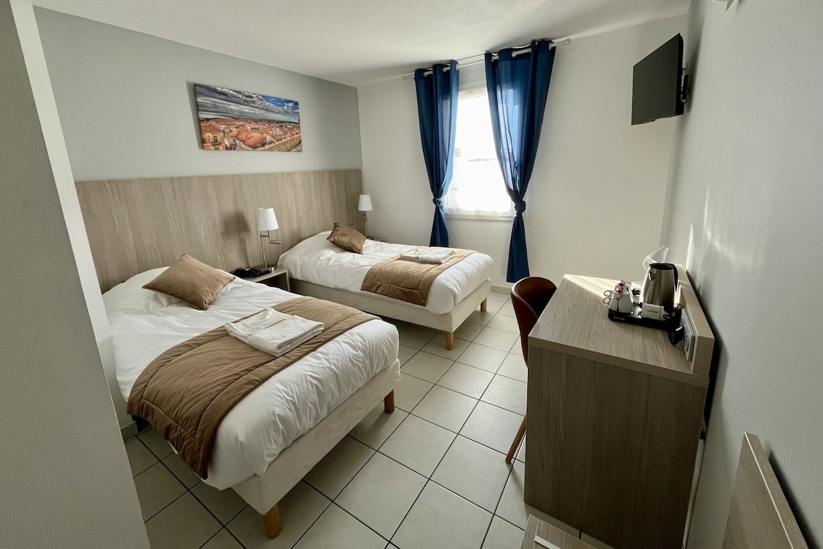Quarto de hotel com duas camas, uma mesa de trabalho com cafeteira e televisão na parede.