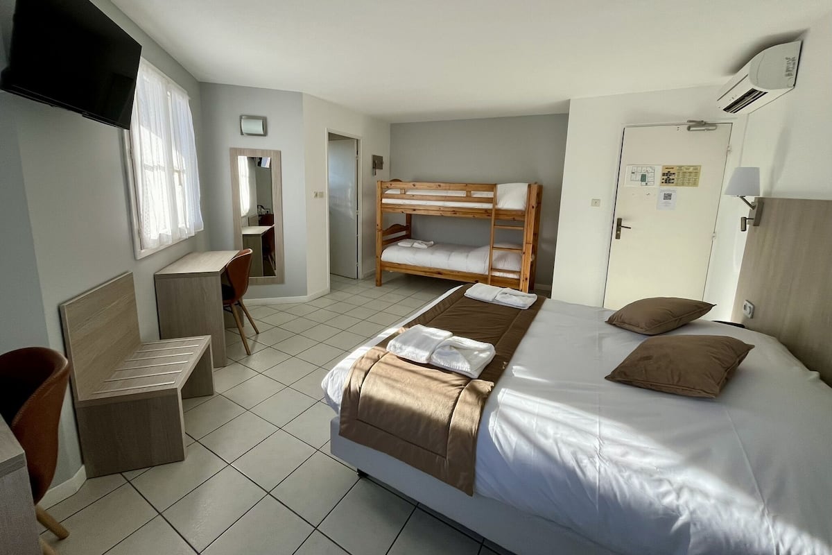 Quarto de hotel com cama, escrivaninha, cadeira, beliche e televisão.
