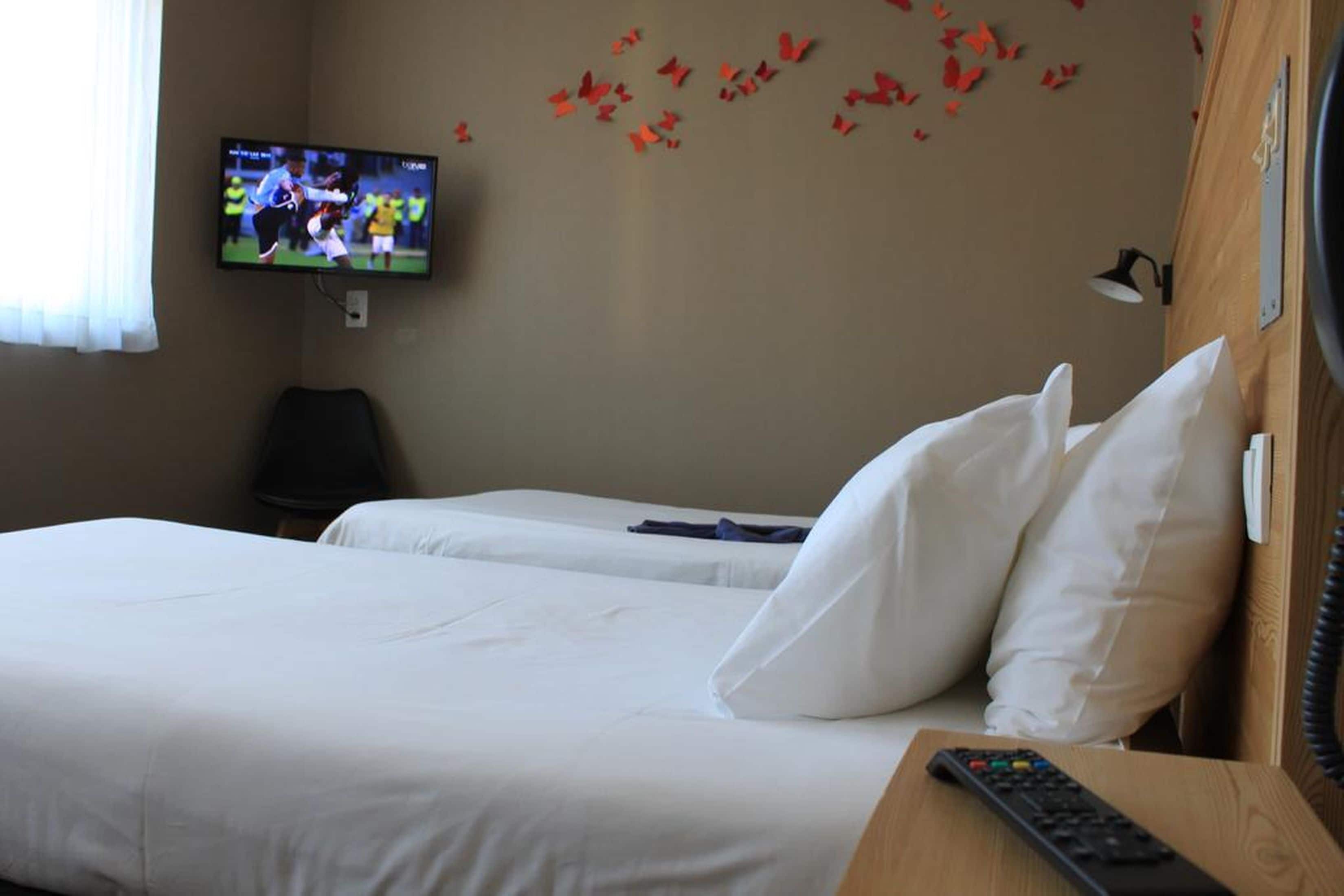 Quarto de hotel com duas camas, uma televisão presa à parede e um controle remoto no criado-mudo.