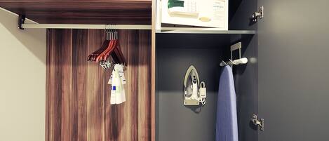 Um guarda-roupa de madeira com espaço para pendurar roupas, um ferro de passar fixado na parede e um porta-toalhas.
