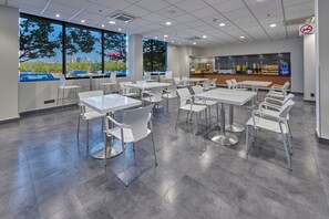 Uma área de refeições moderna com mesas e cadeiras brancas, janelas amplas e bancada de cozinha.