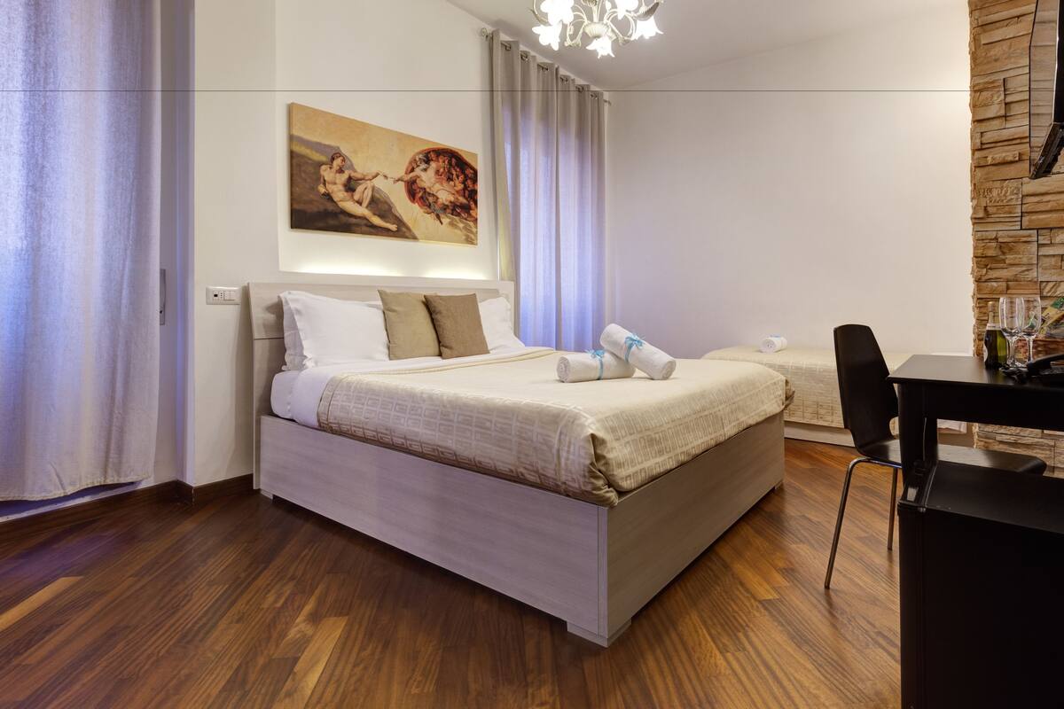 Quarto de hotel moderno com uma cama grande, uma escrivaninha e uma cadeira. Há uma pintura na parede e um candelabro.