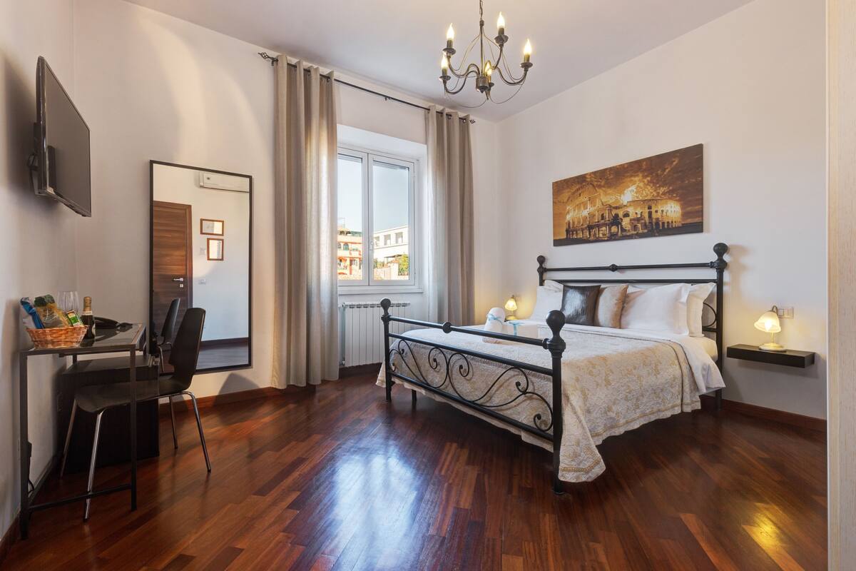 Quarto de hotel com uma cama, uma escrivaninha com cadeira, televisão, janela com cortinas e um lustre.