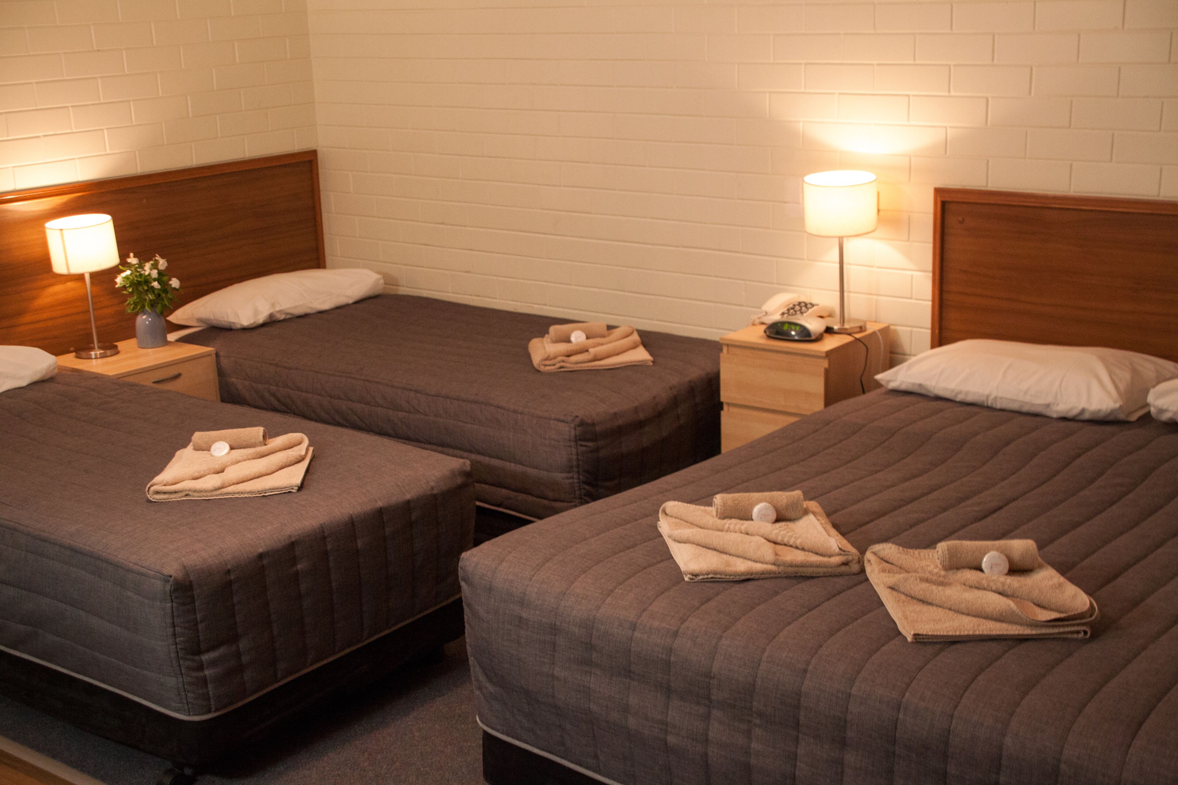 Quarto de hotel com duas camas, cada uma com colcha marrom acolchoada e uma toalha dobrada em cima. Há mesas de cabeceira com luminárias e um telefone.