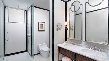 Uma casa de banho moderna com chuveiro envidraçado, sanita branca, lavatório de mármore com duas cubas e um espelho grande sobre a bancada.