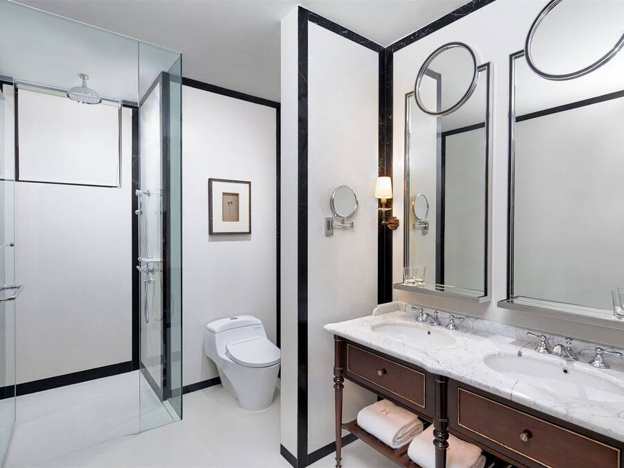 Uma casa de banho moderna com chuveiro envidraçado, sanita branca, lavatório de mármore com duas cubas e um espelho grande sobre a bancada.
