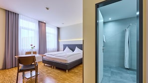 Quarto de hotel com cama, escrivaninha e cadeira. É visível um chuveiro através de uma porta de vidro.