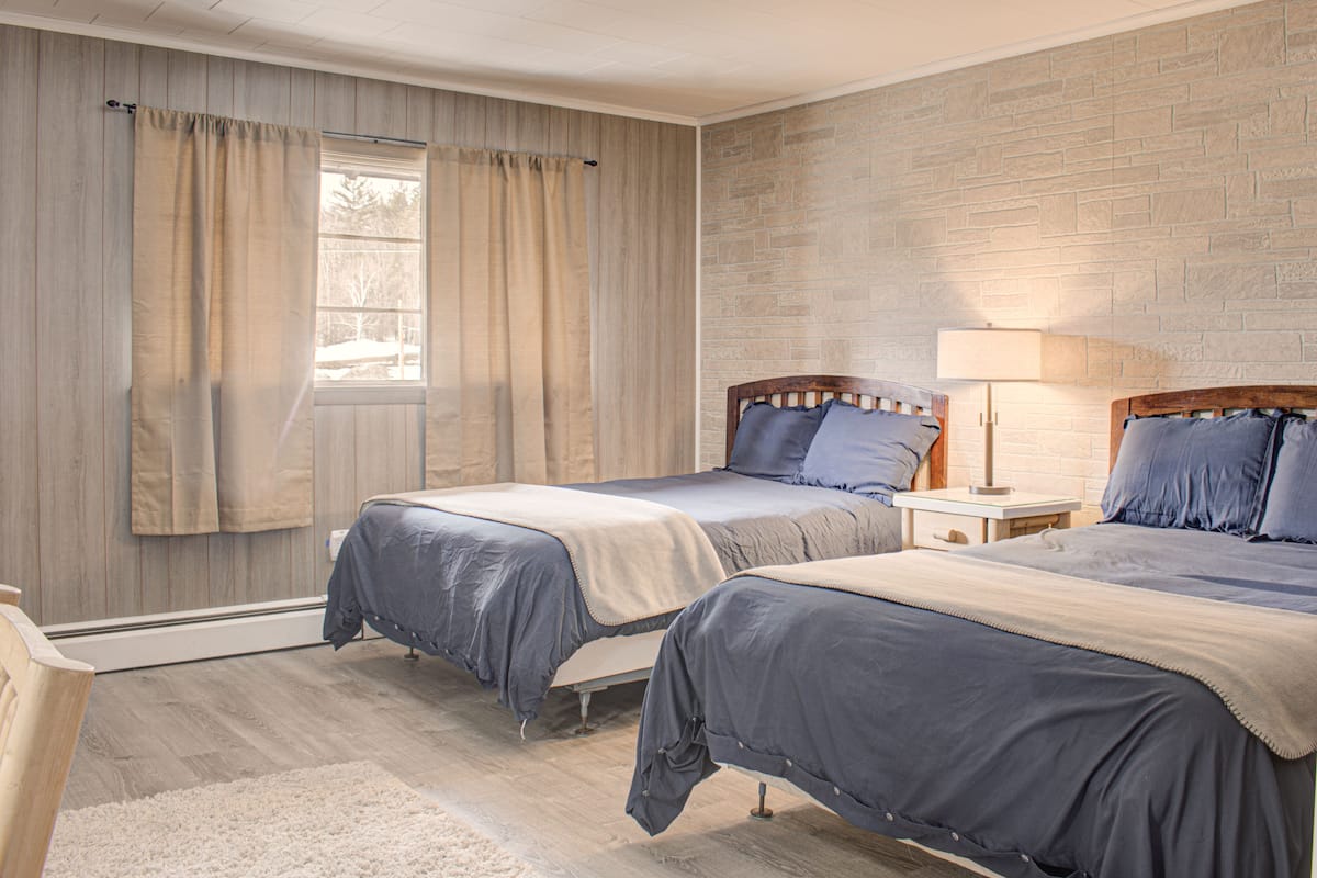 Quarto com duas camas, janela com cortinas, luminária e parede de tijolos.