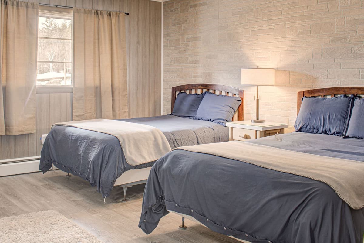 Quarto com duas camas, parede de tijolos, janela com cortinas e abajur.