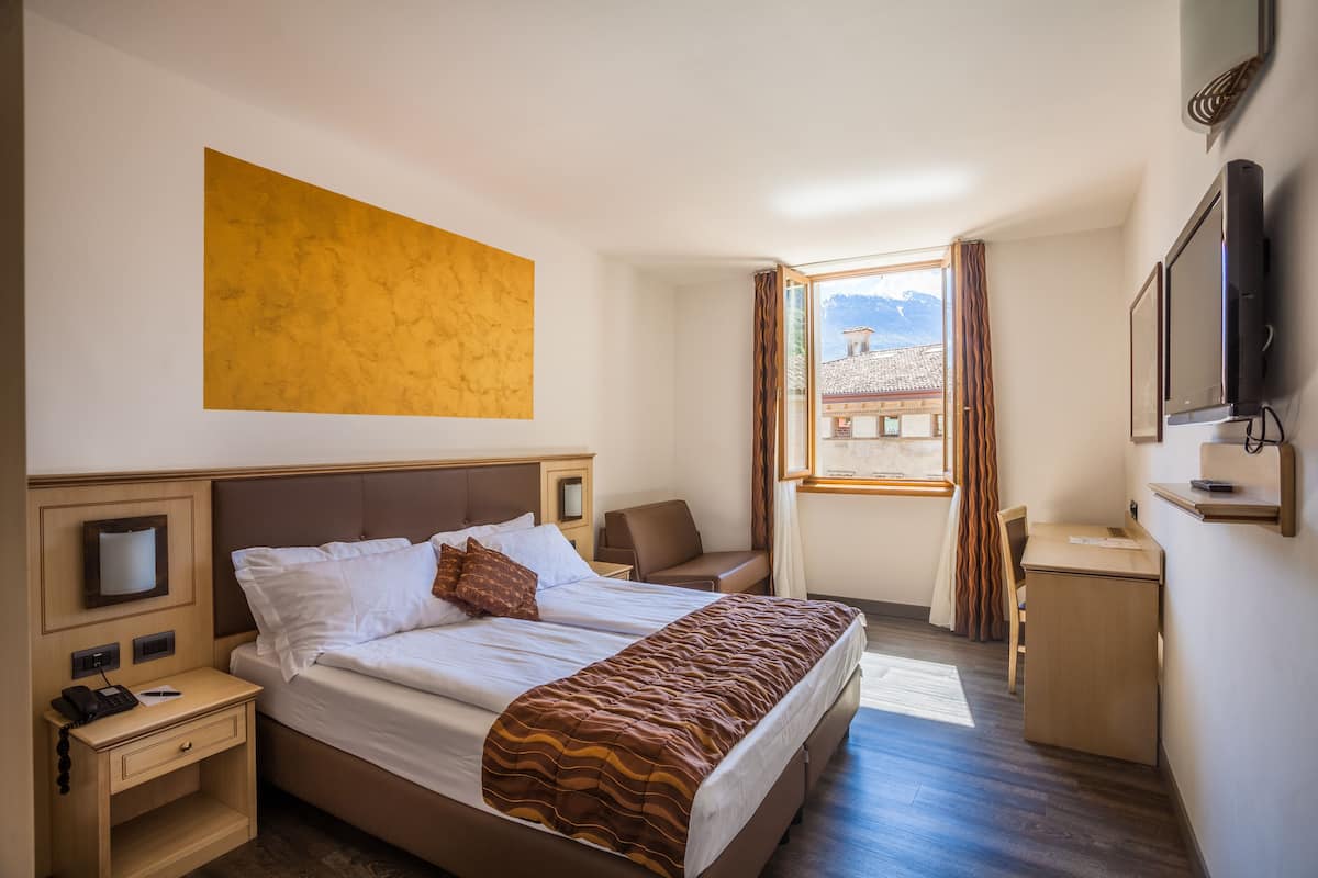 Quarto de hotel com uma cama, escrivaninha, cadeira, televisão e uma grande janela com cortinas.
