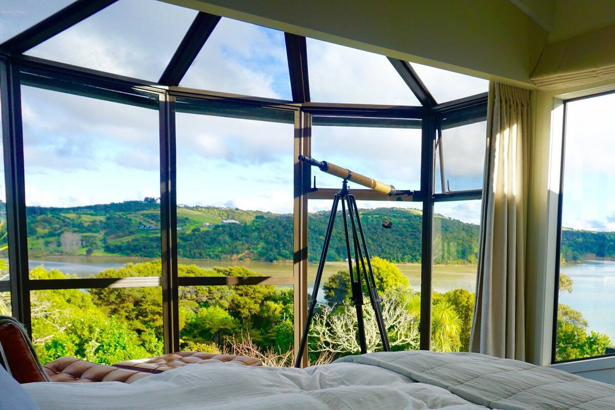 Quarto com uma janela grande que oferece vista para um rio e colinas.