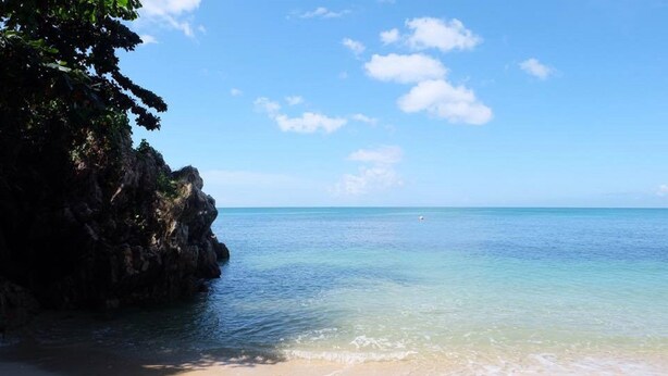 Uma praia com águas turquesa cristalinas, areal arenoso e falésias rochosas.