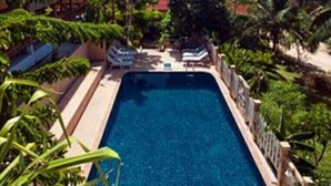 Uma piscina retangular rodeada por espreguiçadeiras e palmeiras, com vista para o oceano e um prédio com telhado vermelho ao fundo.