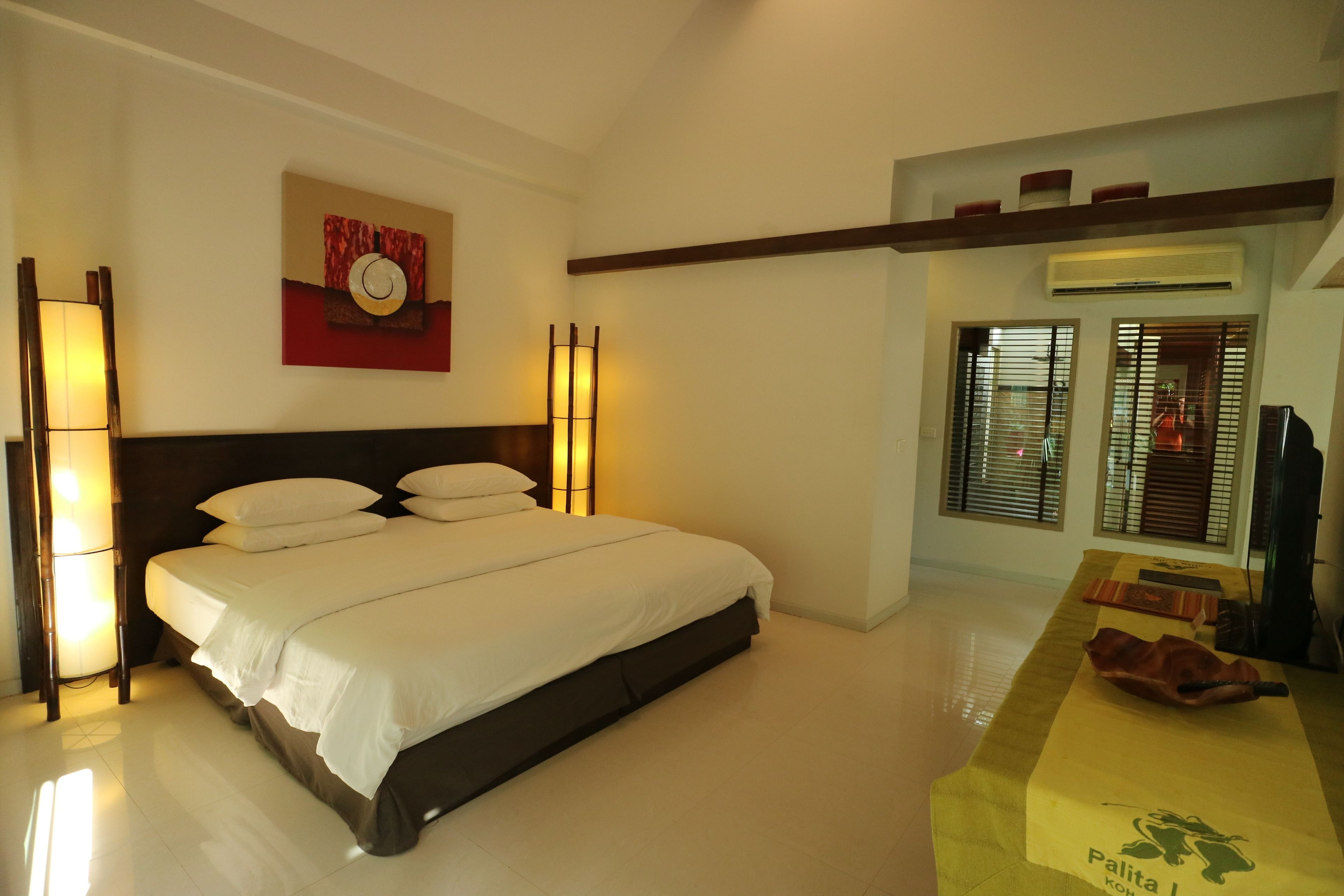 Quarto de hotel com uma cama grande, abajures, um relógio na parede e uma janela com persianas.