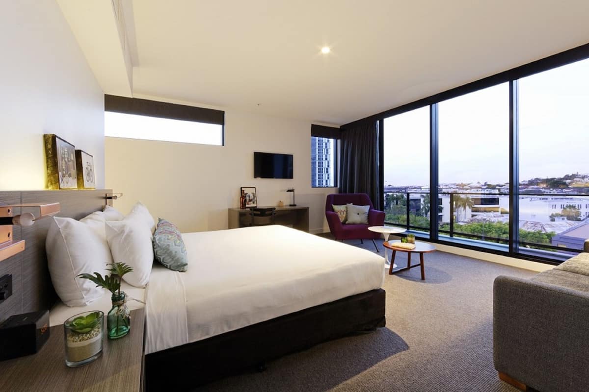 Quarto de hotel moderno com uma cama grande, uma escrivaninha, uma cadeira e vista para a paisagem urbana através de amplas janelas.