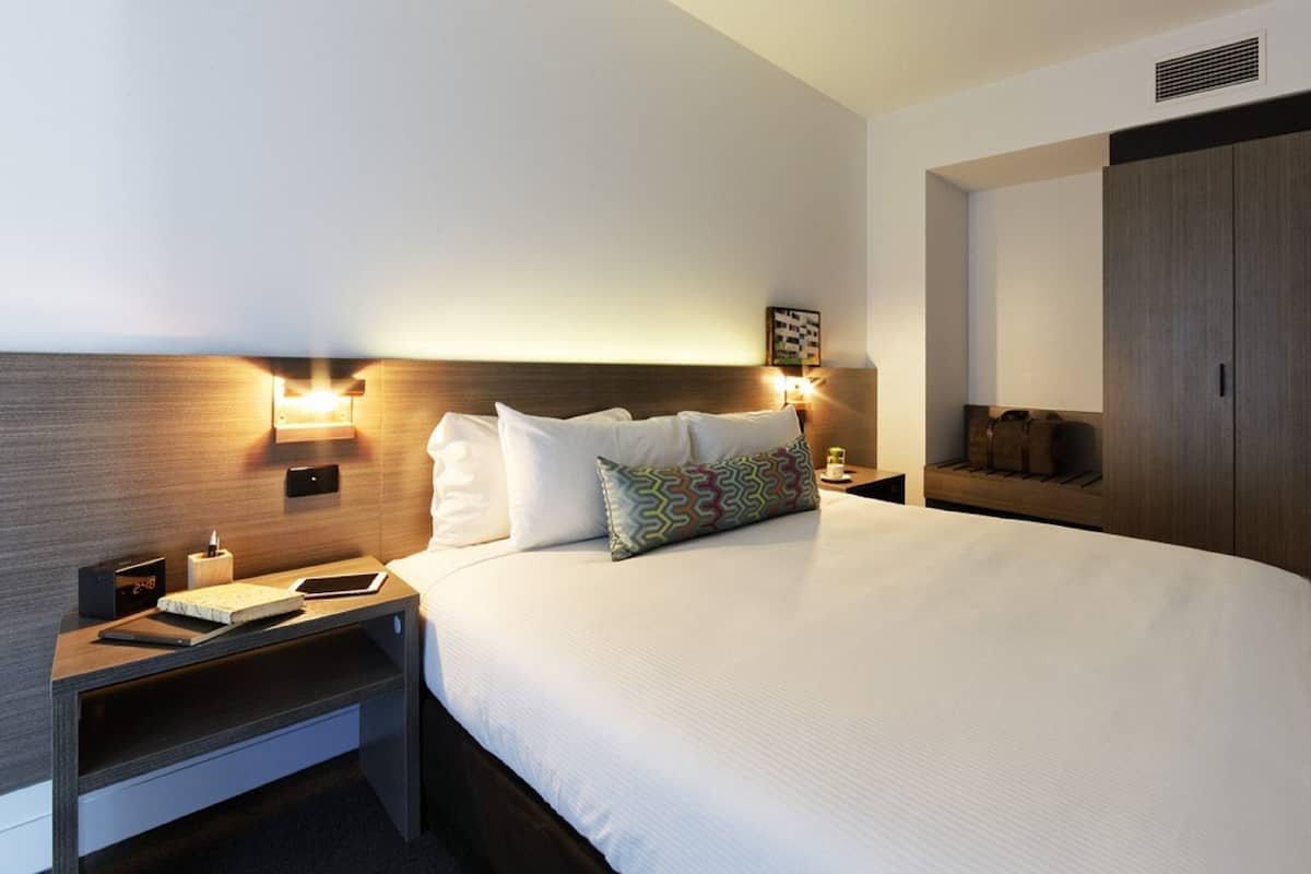 Quarto de hotel com uma cama grande, mesas de cabeceira, um guarda-roupa e luzes fixadas na parede.