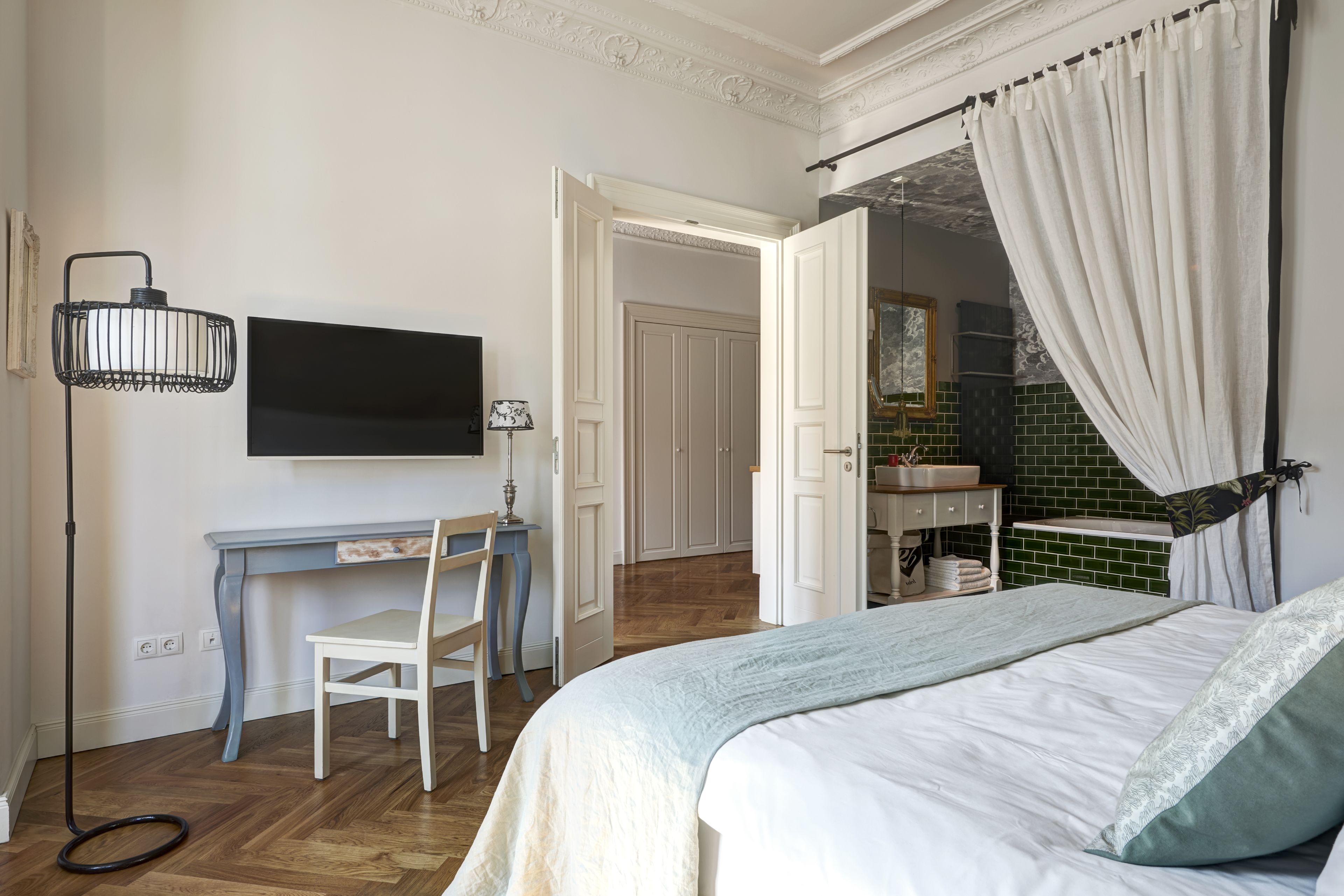 Quarto com cama, escrivaninha, cadeira, televisão e banheiro visível por uma porta aberta.