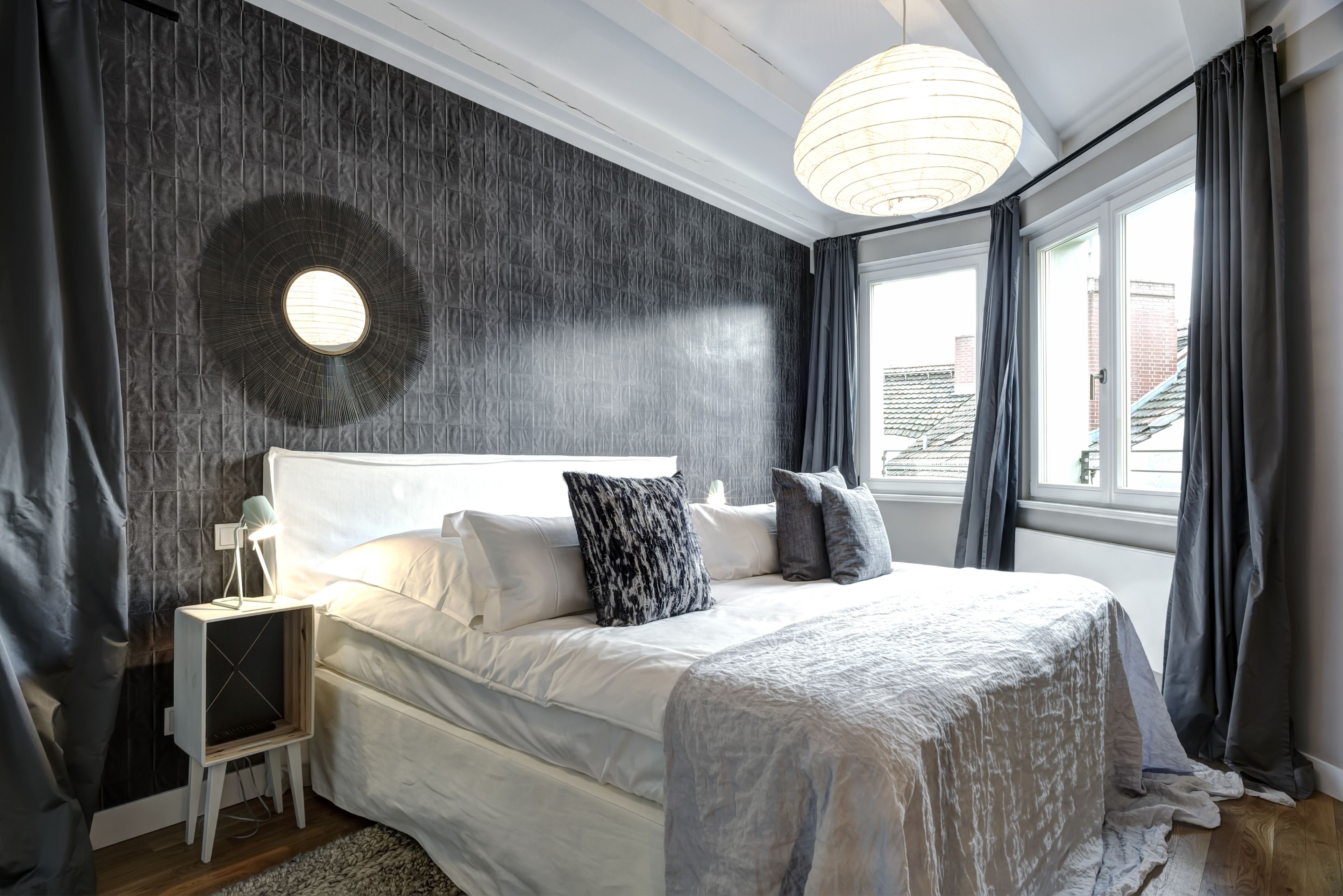 Quarto moderno com uma cama grande, uma mesinha de cabeceira com abajur, uma luminária circular na parede e uma janela com cortinas.